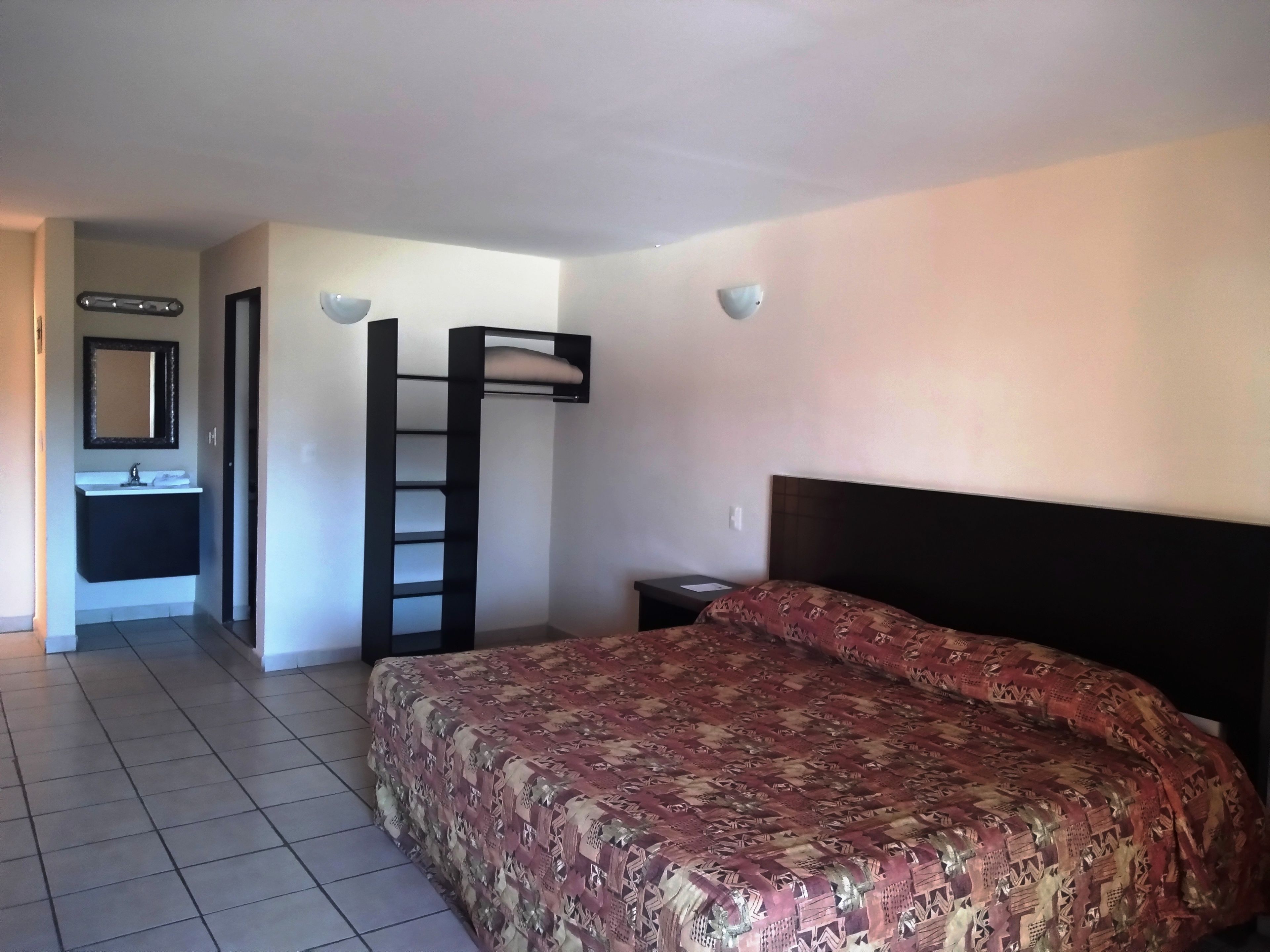 Quarto com cama, prateleira embutida, mesa de cabeceira e banheiro com pia e espelho.