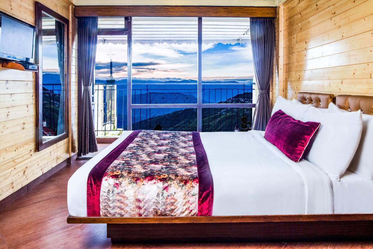 Quarto com cama grande, vista para montanhas e um lago, paredes de madeira e uma varanda.