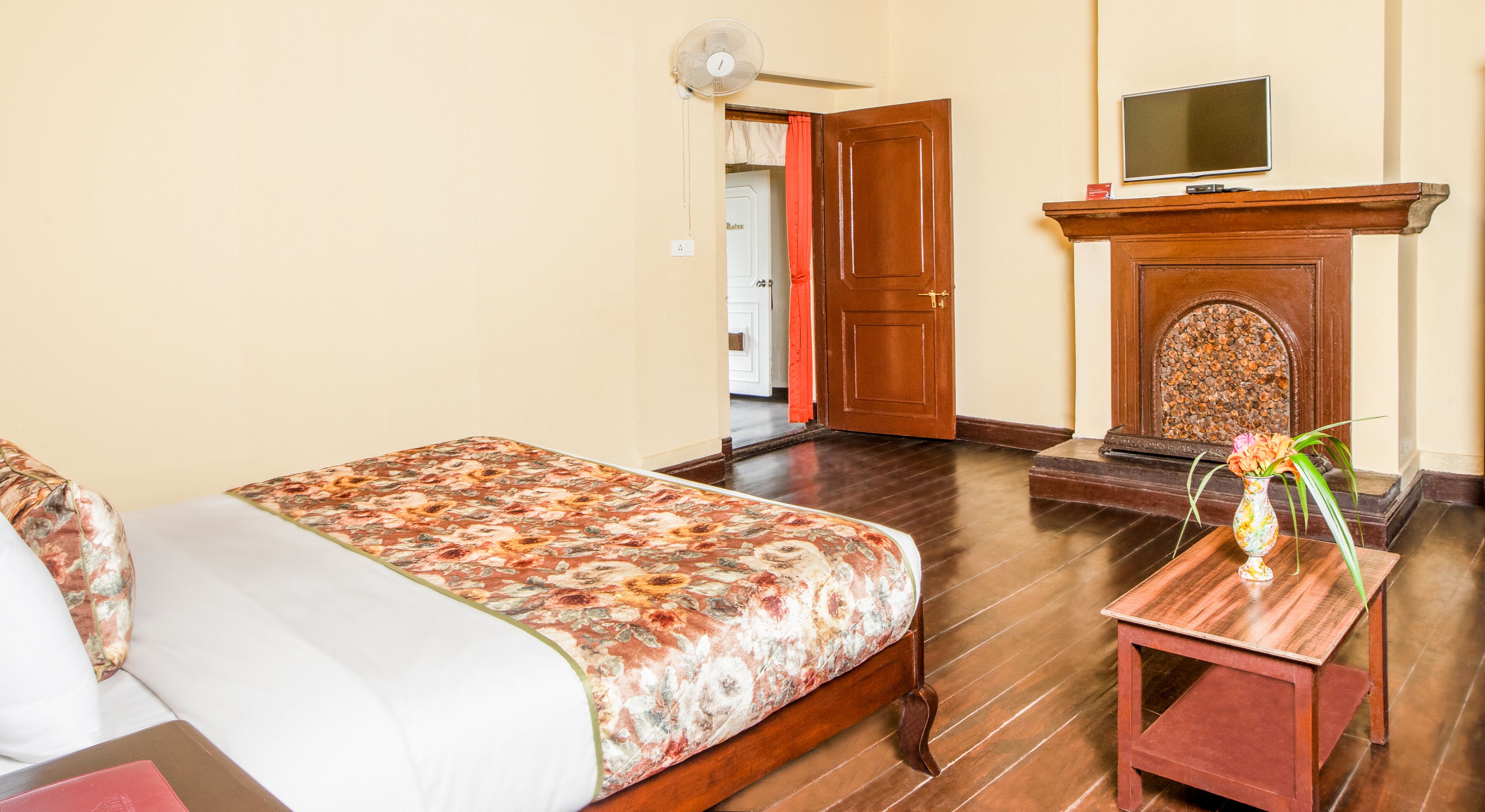 Quarto de hotel com cama, piso de madeira, TV sobre lareira e uma pequena mesa com um vaso de flores.