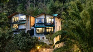 Uma casa moderna de dois andares com varandas, rodeada por uma floresta densa e vista para uma colina.