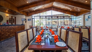 Restaurante com teto de madeira e janelas amplas. As mesas estão dispostas com guardanapos laranja e talheres.