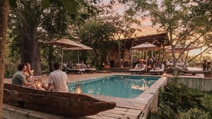 Uma área à beira da piscina com espreguiçadeiras e guarda-sóis, cercada por árvores e um deck de madeira.