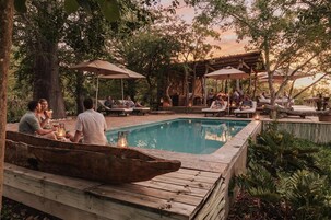 Uma área à beira da piscina com espreguiçadeiras e guarda-sóis, cercada por árvores e um deck de madeira.