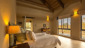 Quarto com cama grande, mesa de cabeceira, luminária, pequena mesa e vista para o exterior.