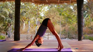 Uma pessoa praticando uma postura de yoga em um tapete roxo, em ambiente externo com árvores e uma estrutura de telhado de palha.