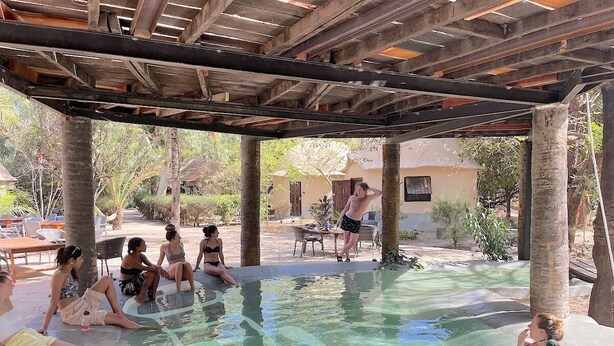 Uma área de piscina com água cristalina, vigas de madeira e palmeiras.