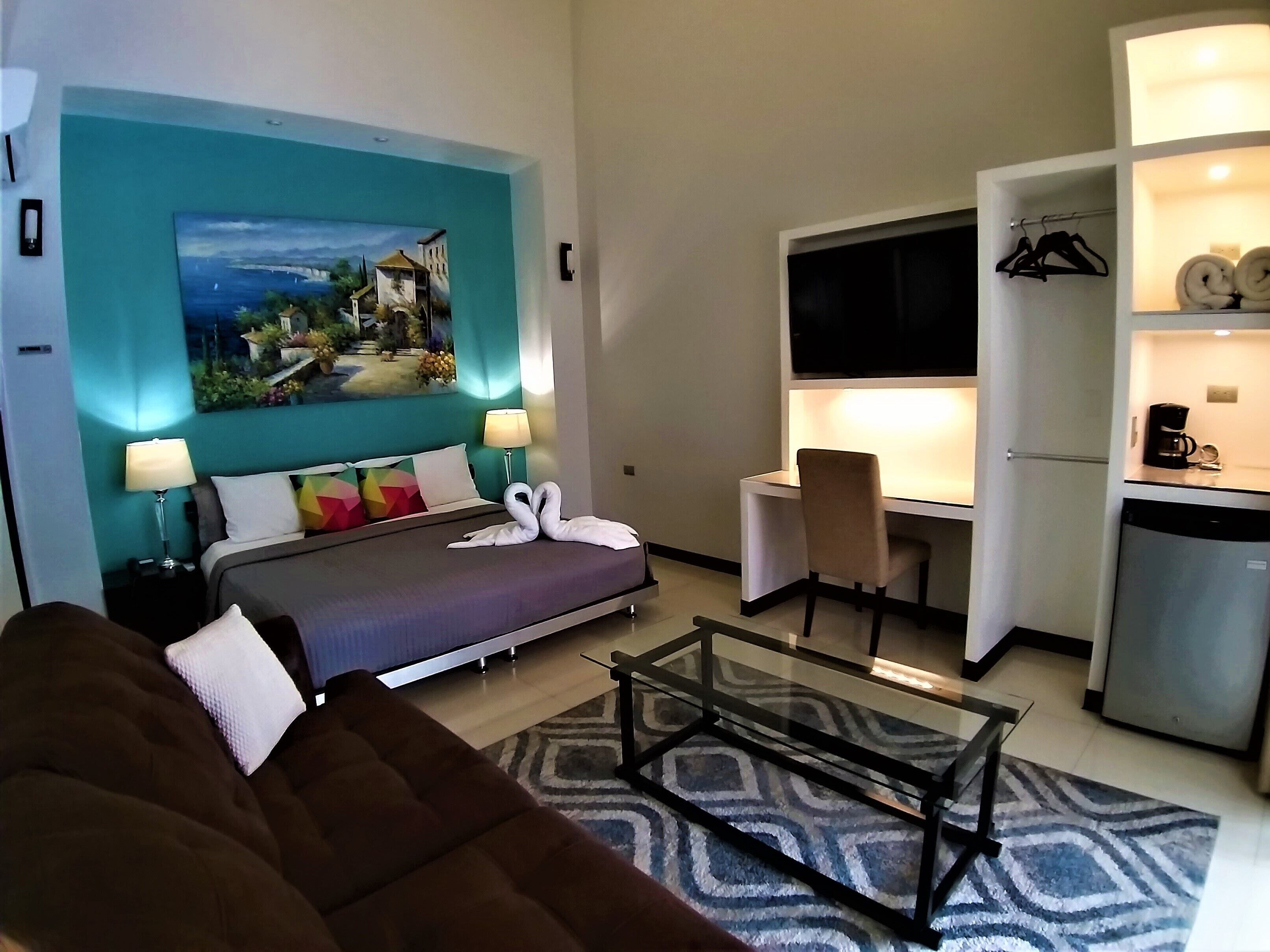 Quarto de hotel com cama, escrivaninha, poltrona, sofá, televisão e cozinha.