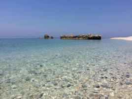 Uma praia clara com águas rasas e pedras, um afloramento rochoso e uma área arenosa.
