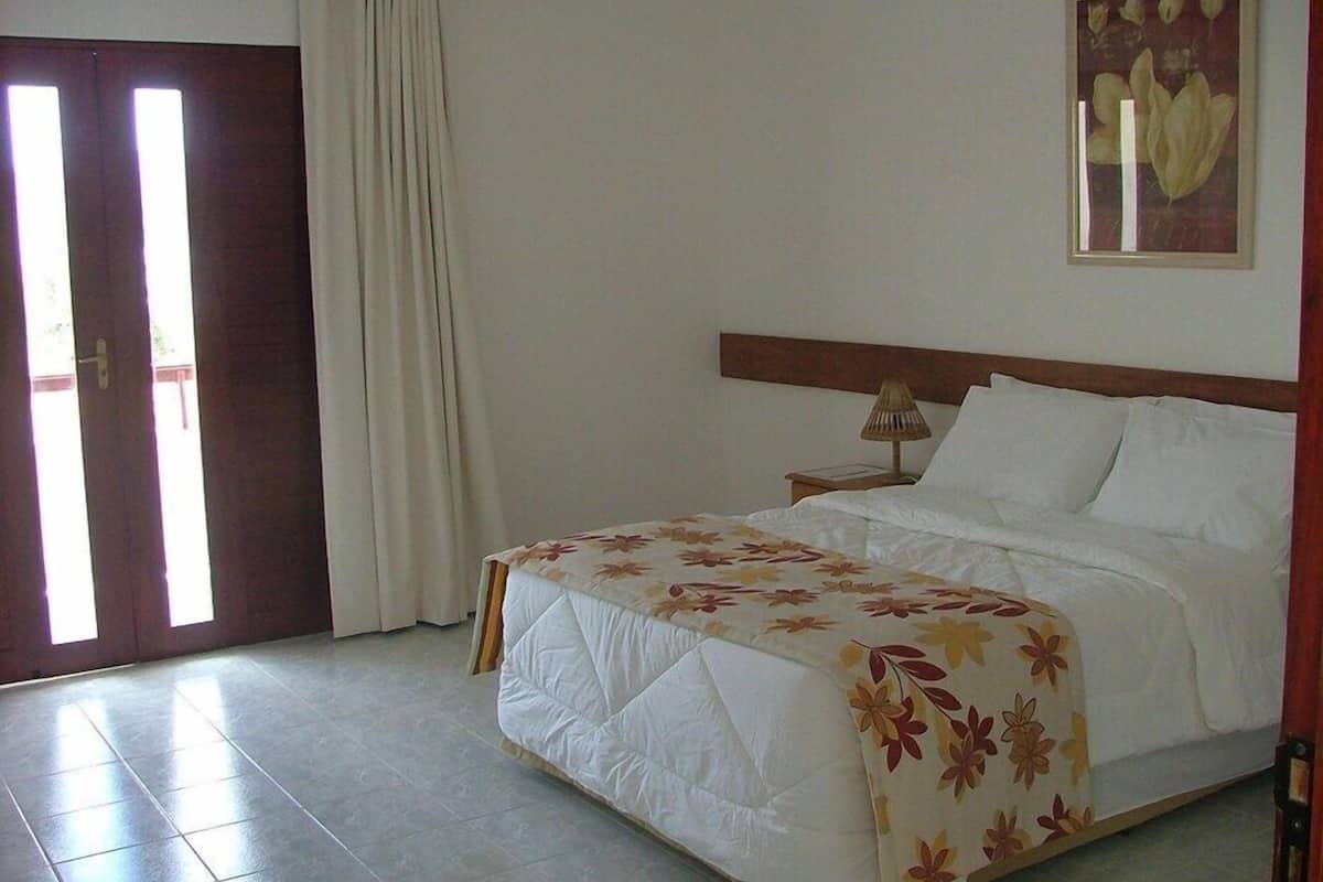 Quarto com cama, mesa de cabeceira, luminária, porta com cortinas e um quadro na parede.