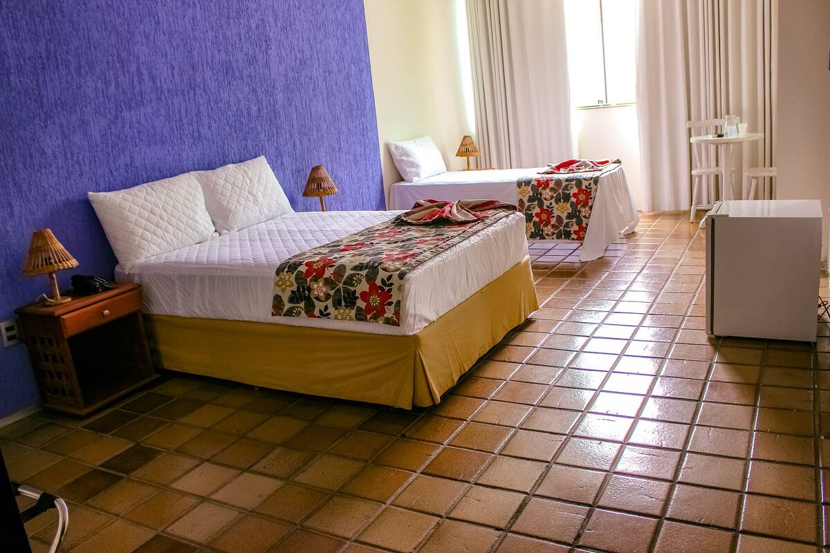 Quarto de hotel com cama, mesa de cabeceira, abajur e uma pequena mesa com cadeira.