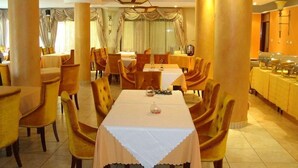 Uma área de refeições com mesas e cadeiras redondas, um candelabro e janelas amplas com cortinas.