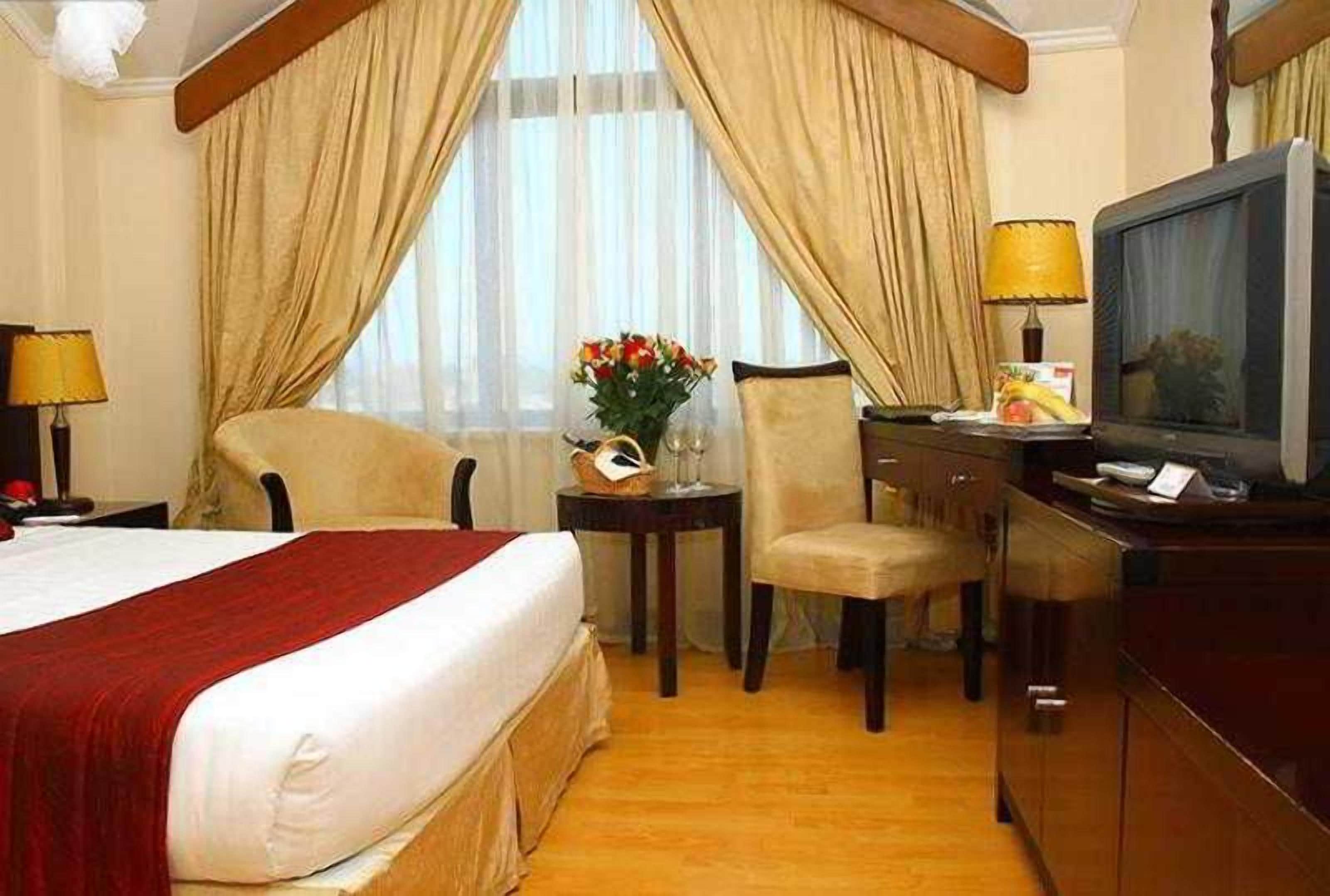 Quarto de hotel com cama, escrivaninha com televisão, cadeira e pequena mesa com um vaso de flores.