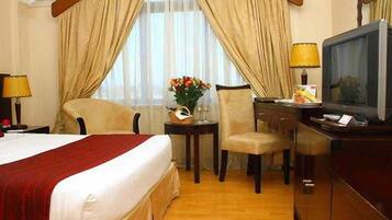 Quarto de hotel com cama, escrivaninha com televisão, cadeira e pequena mesa com um vaso de flores.