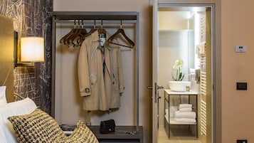Quarto de hotel com cama, mesa de cabeceira com abajur, guarda-roupa com roupas penduradas e banheiro visível por uma porta aberta.