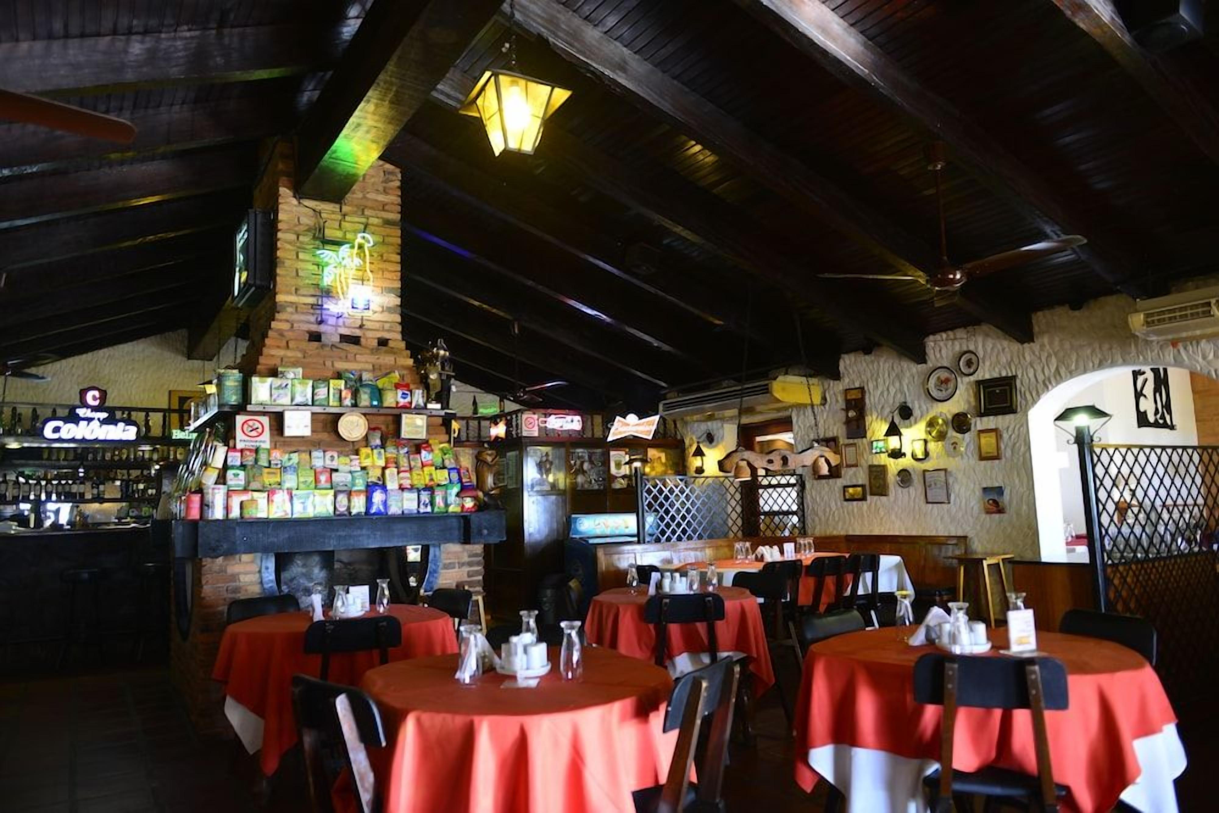 Interior de restaurante com várias mesas, um espaço de bar e uma parede de tijolos com diversos objetos expostos.
