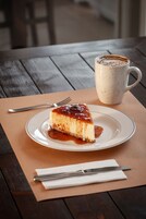 Uma fatia de cheesecake em um prato com garfo e faca, uma xícara de café e um guardanapo sobre uma mesa de madeira.