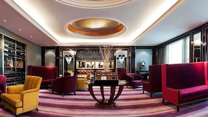 Um lobby luxuoso com uma mesa redonda central, assentos vermelhos e uma adega visível através de um painel de vidro.