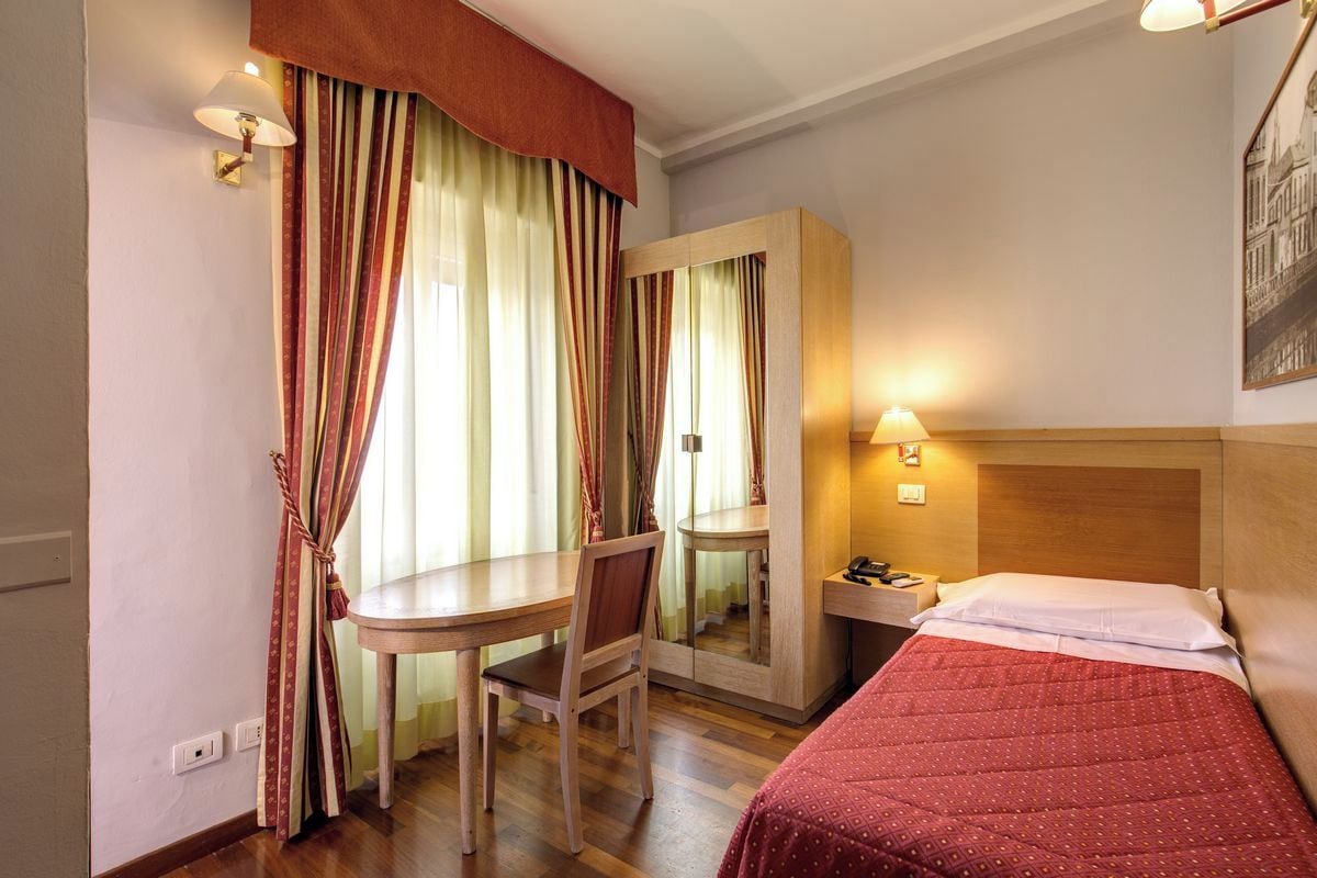 Quarto de hotel com cama, escrivaninha, cadeira, espelho e mesa de cabeceira.