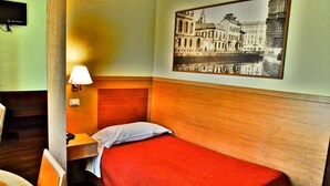 Uma cama de solteiro com um cobertor vermelho, cabeceira de madeira, mesa de cabeceira com abajur e um quadro com uma foto de um edifício na parede.