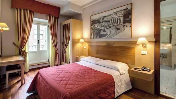 Quarto de hotel com cama, mesas de cabeceira, escrivaninha, cadeira, janela com cortinas, luminária na parede e um quadro emoldurado.