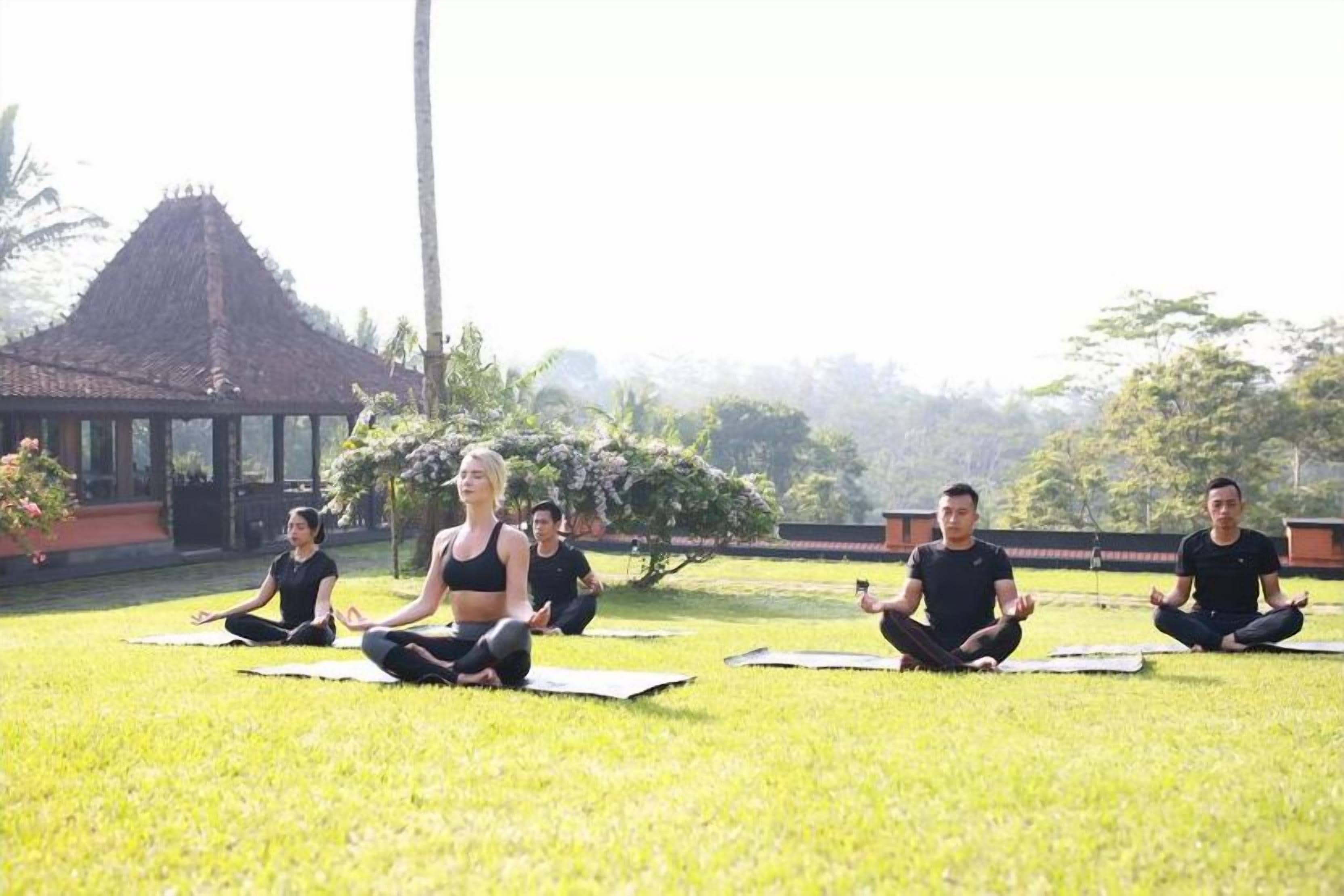 Um grupo de pessoas meditando na grama em ambiente externo, com um edifício tradicional ao fundo.