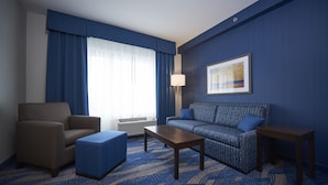Quarto de hotel com uma poltrona marrom, um sofá azul, uma mesa de centro de madeira e um banquinho azul. Há uma janela grande com cortinas azuis, um abajur e um quadro na parede.