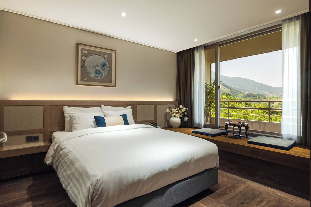 Quarto de hotel moderno com uma cama grande, uma mesa de cabeceira, uma janela com vista para as montanhas e um quadro na parede.