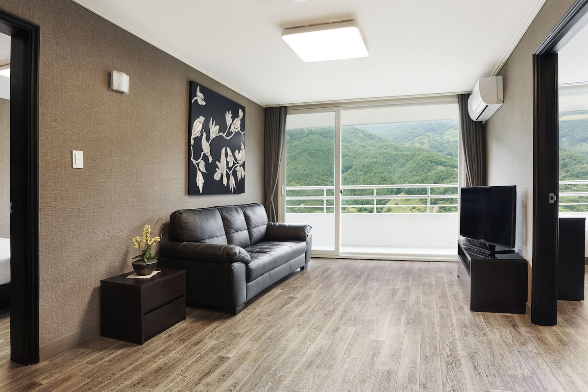 Uma sala de estar moderna com um sofá de couro, uma TV de tela plana e uma grande janela com vista para colinas verdes.