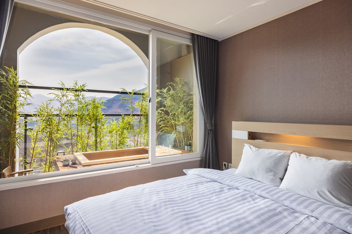 Quarto de hotel com uma grande janela, cama com roupa de cama branca e vista para bambus e montanhas.