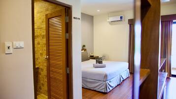 Quarto de hotel com porta de madeira, cama com colcha branca, mesa de cabeceira com abajur e ar condicionado na parede.