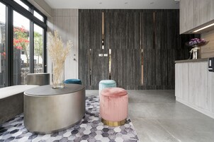 Um lobby moderno com uma mesa metálica redonda, um banquinho rosa e um apoio para os pés azul. Uma grande porta de madeira escura com fendas verticais é visível ao fundo.