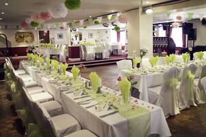 Salão de banquetes com longas mesas montadas para um evento formal, com toalhas brancas e guardanapos verdes.