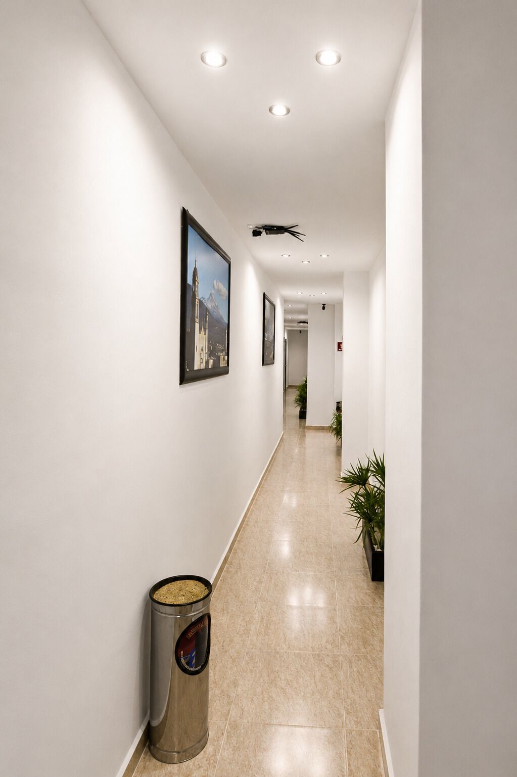 Um corredor com um cesto de lixo, um projetor no teto e quadros nas paredes.