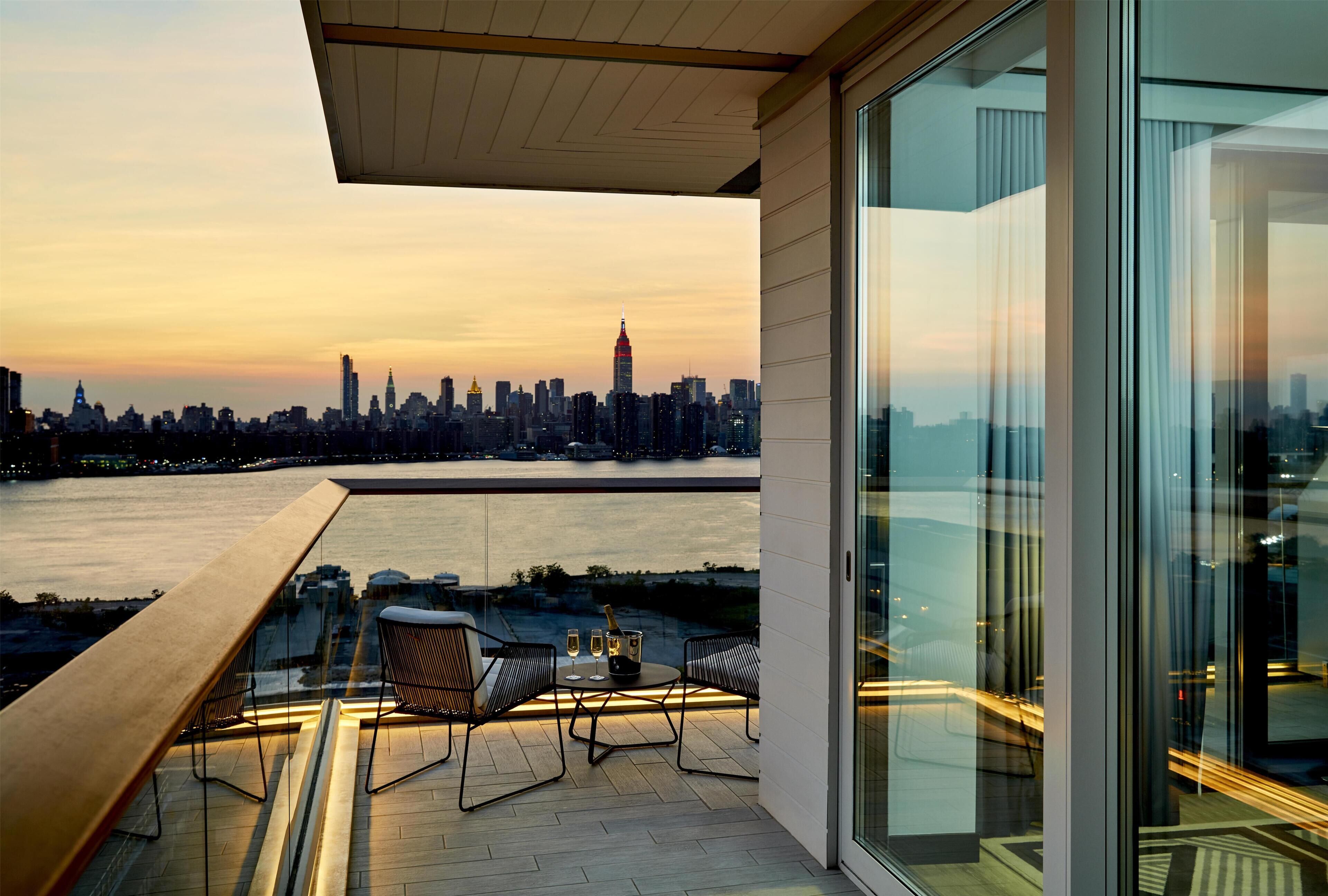 Uma varanda com mesa e cadeiras, com vista para o horizonte urbano e um corpo de água.
