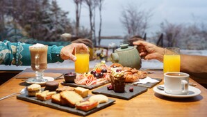 Mesa de madeira com diversas comidas e bebidas, com vista para um corpo d'água.