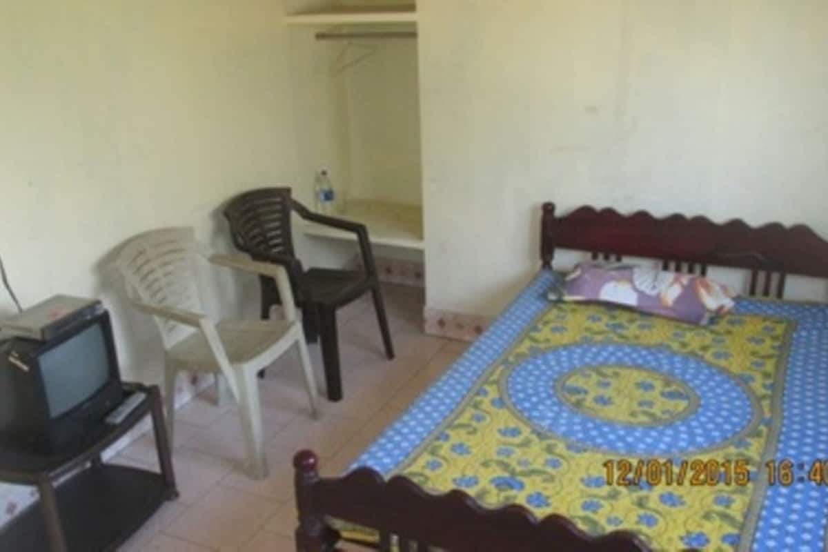 Quarto pequeno com uma cama, uma televisão sobre um suporte e duas cadeiras.