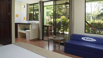 Quarto de hotel com uma cama, uma pequena mesa com um cesto, um sofá azul com o logotipo 'MAVI', uma cozinha compacta e uma porta de vidro deslizante que dá para uma varanda com plantas e uma rede.