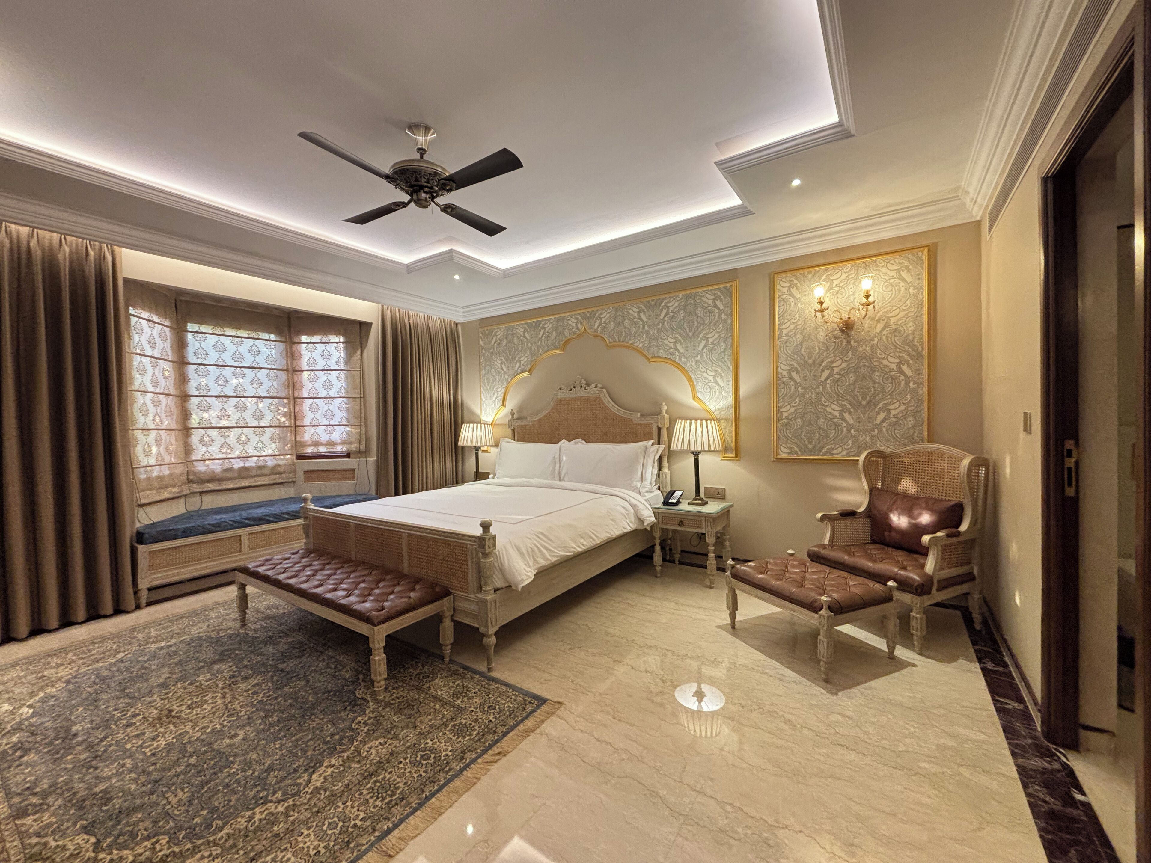 Um quarto espaçoso com uma cama grande, ventilador de teto, área de estar com uma poltrona e um detalhe decorativo na parede.