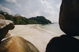 Uma praia com pedras grandes em primeiro plano, um barco na água e uma ilha coberta por floresta ao fundo.