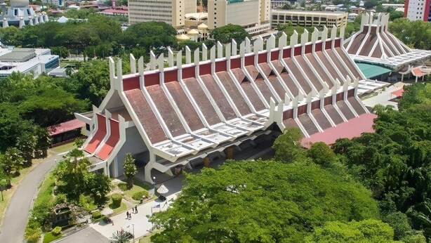 Um amplo anfiteatro moderno com assentos em níveis escalonados e palco central, cercado por vegetação exuberante e edifícios urbanos.