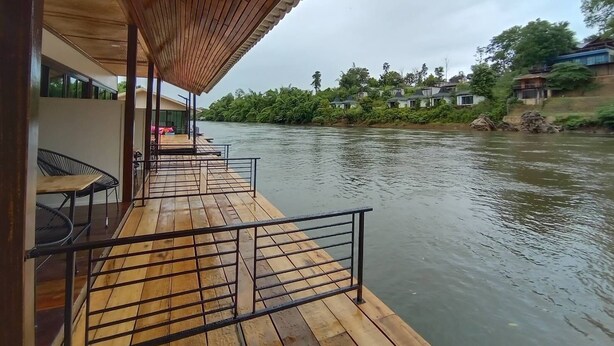Um deck de madeira com grades de metal dando vista para um rio, com edifícios e vegetação ao fundo.