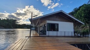 Um píer de madeira se estende sobre um rio calmo, conduzindo a uma casa moderna com telhado inclinado e grandes janelas.