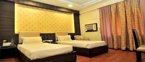 Quarto de hotel com duas camas, uma mesa de cabeceira, uma cadeira, uma janela grande com cortinas e um ar condicionado de parede.
