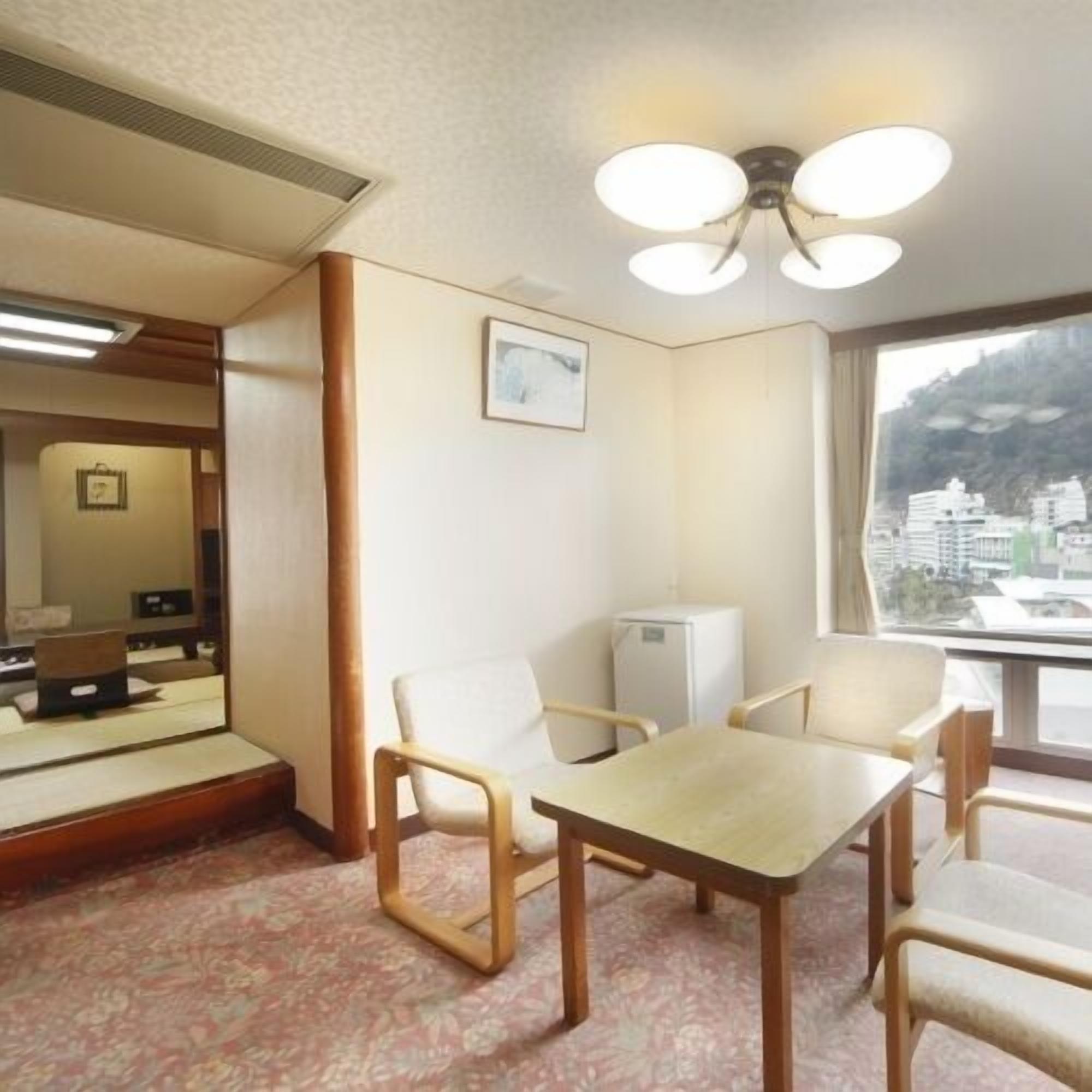 Quarto com uma mesa de madeira, duas cadeiras, um pequeno frigorífico branco, um quadro na parede e uma janela com vista para edifícios e colinas.