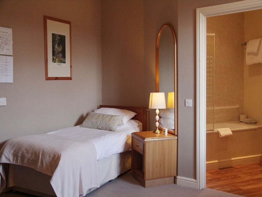 Quarto de hotel com cama, mesa de cabeceira com luminária, espelho e chuveiro.