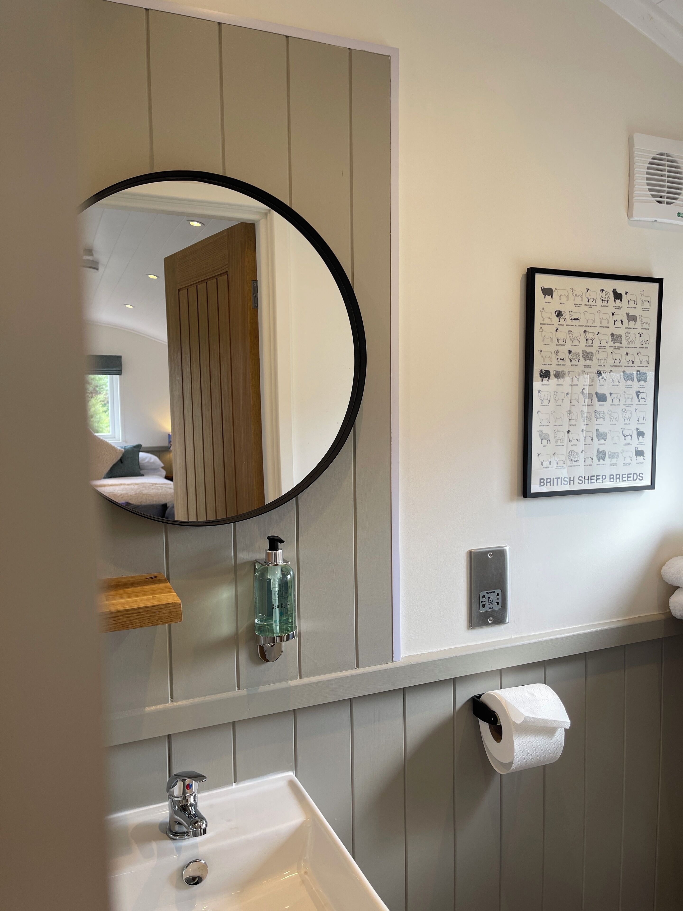 Casa de banho com espelho, quadro emoldurado com raças de ovelhas britânicas, dispensador de sabonete, porta-toalhas e suporte de papel higiénico.