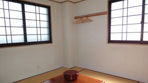 Um quarto com piso de tatame, uma mesa de madeira com uma tigela de madeira escura, e duas janelas com molduras pretas.