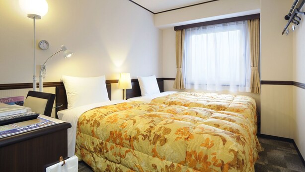 Quarto de hotel com duas camas, uma escrivaninha com abajur, relógio e janela com cortinas.