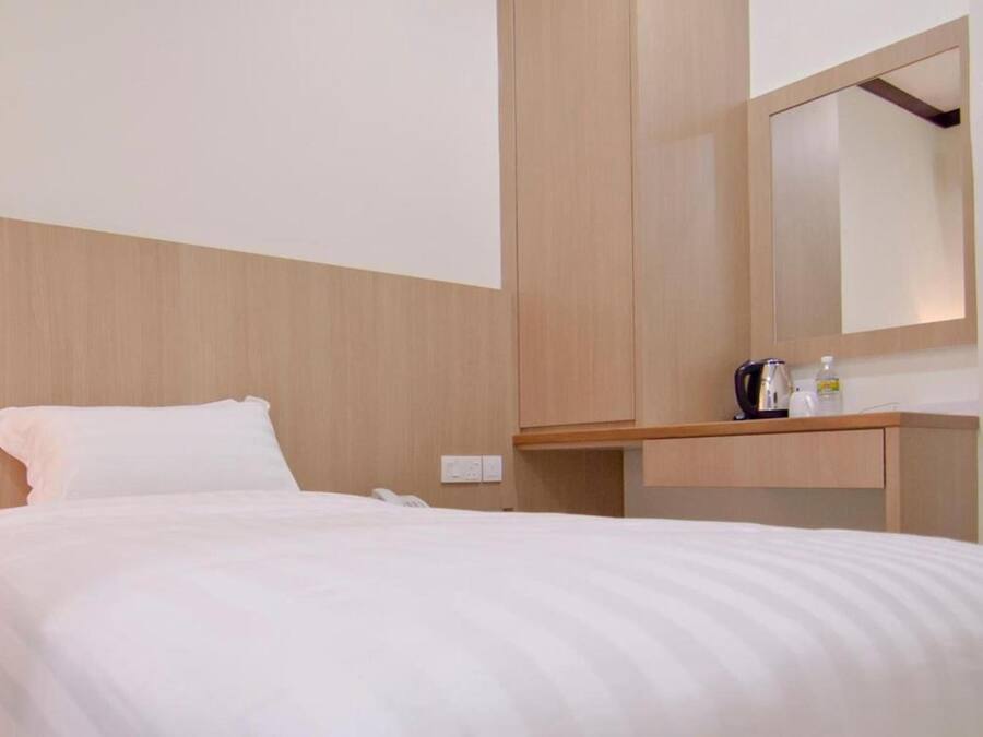 Quarto de hotel com cama, mesa de cabeceira, luminária na parede, espelho e prateleira com artigos de higiene pessoal.