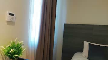 Quarto de hotel com cama, cabeceira, janela com cortinas, termostato fixado na parede e um vaso de planta.